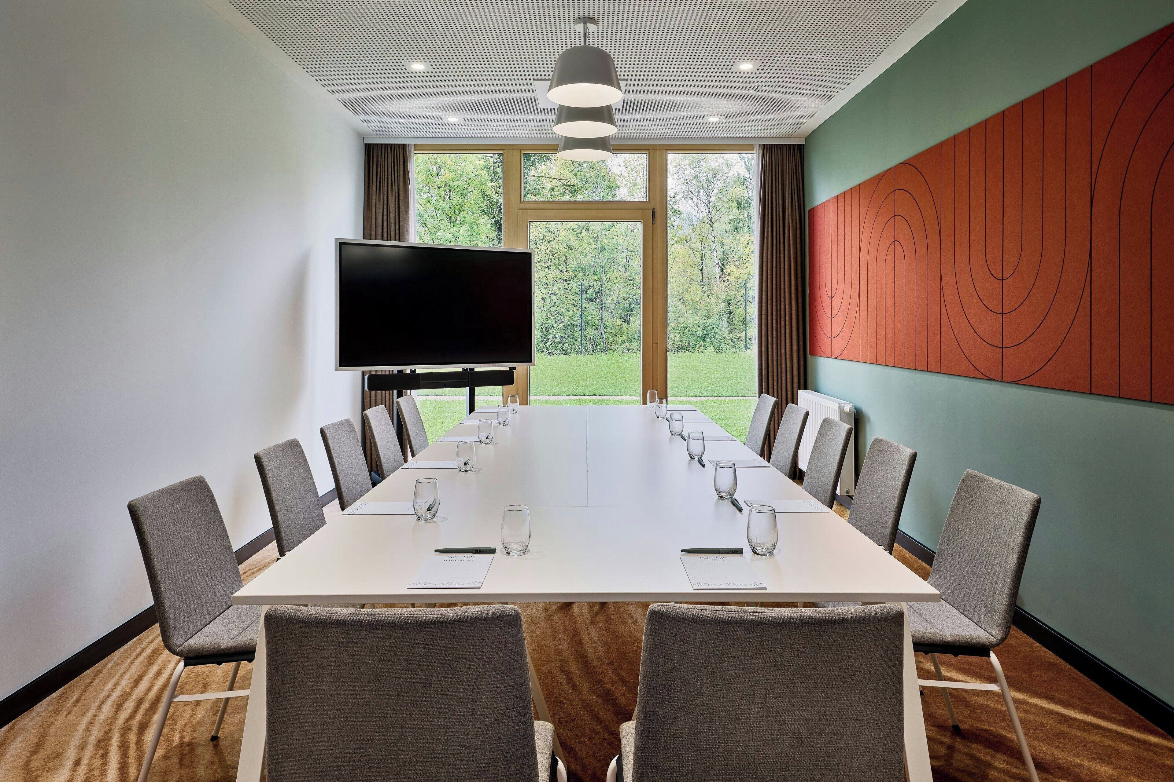 Uma sala de conferências com uma mesa comprida, cadeiras, uma TV de tela plana e vista para o verde através de grandes janelas.