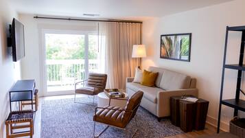 Uma sala de estar com sofá, duas poltronas, mesa de centro, televisão e varanda com cortinas.
