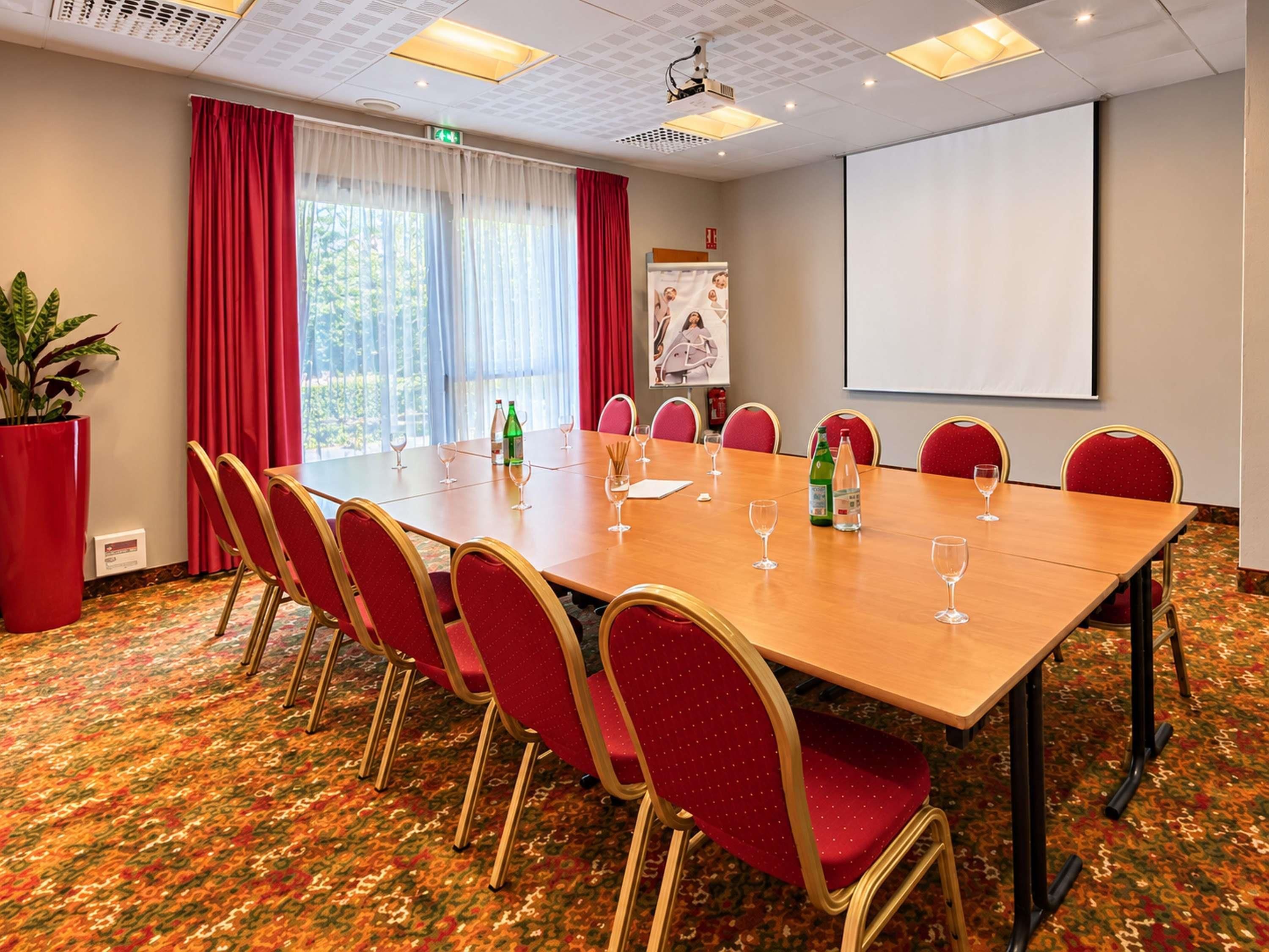 Uma sala de conferências com uma mesa comprida, cadeiras vermelhas, uma tela de projetor e uma janela grande com cortinas.