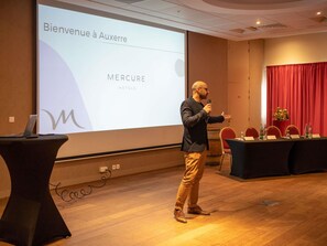 Apresentação em sala de conferências, com uma tela exibindo 'Bienvenue à Auxerre' e 'MERCURE HOTELS'.