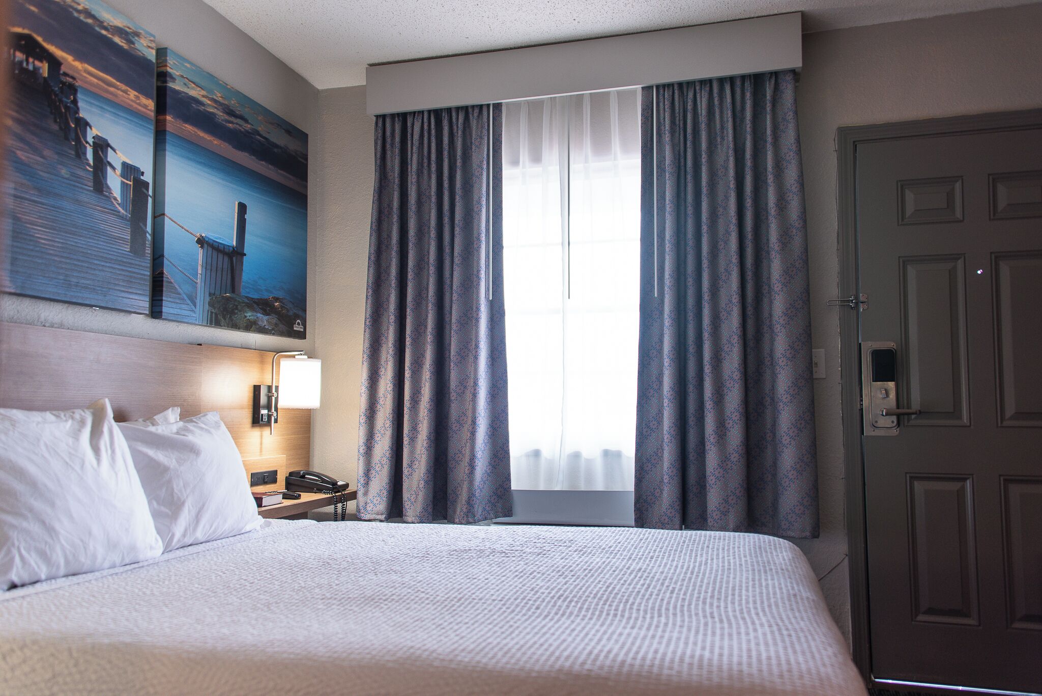 Quarto de hotel com uma cama grande, mesa de cabeceira com abajur, pintura na parede e janela com cortinas.