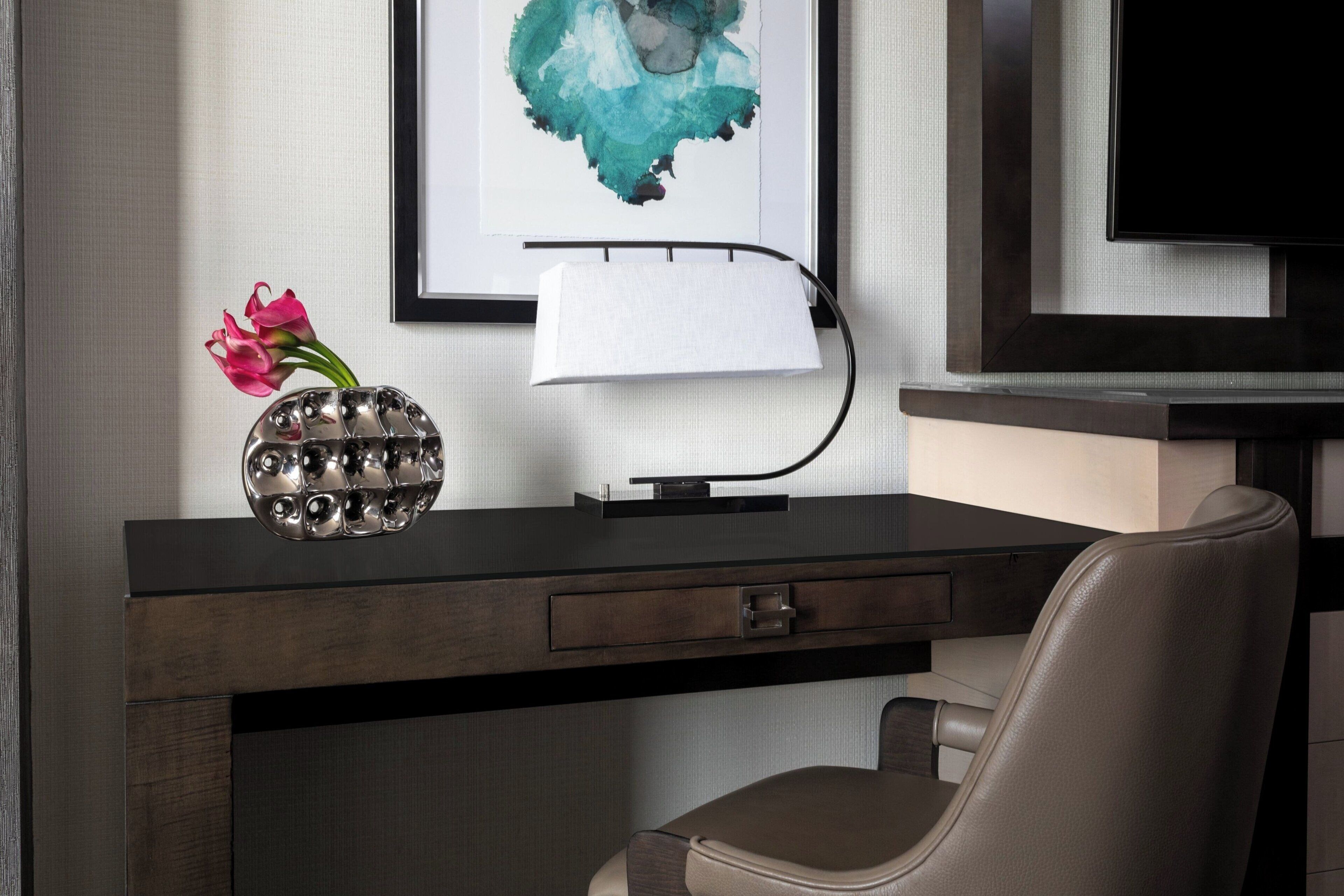 Quarto de hotel moderno com escrivaninha de madeira escura, abajur branco, pintura abstrata emoldurada e um vaso com flores cor-de-rosa.