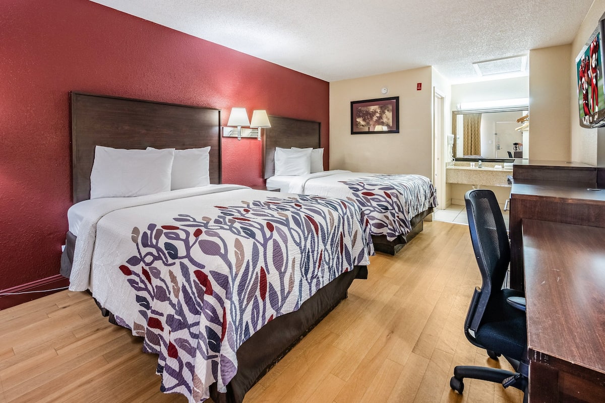 Quarto de hotel com duas camas, uma escrivaninha, uma cadeira e um banheiro visível por uma porta aberta.