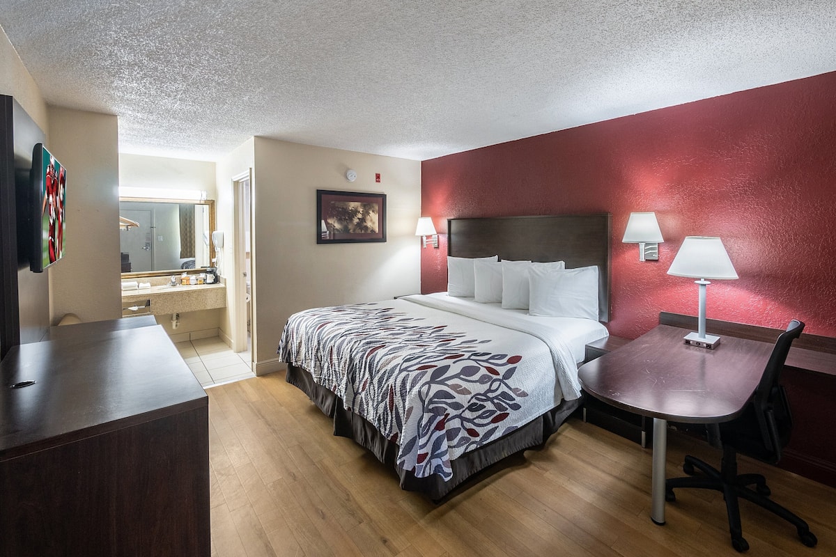 Quarto de hotel com cama, escrivaninha e cadeira, espelho e televisão.