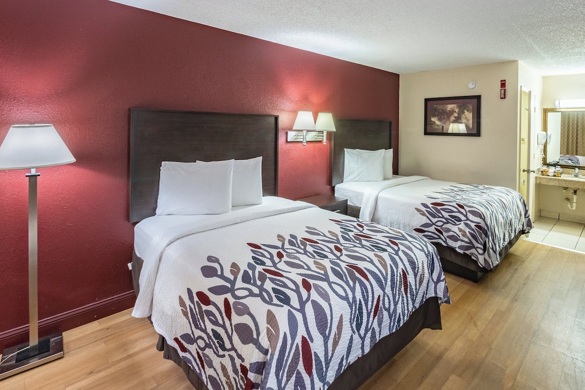 Quarto de hotel com duas camas, uma mesa de cabeceira, uma luminária e banheiro com pia e espelho.