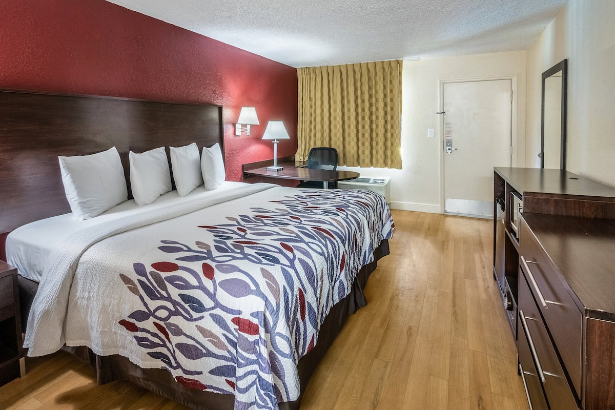 Quarto de hotel com uma cama grande, uma escrivaninha com cadeira, luminária de mesa e cômoda.