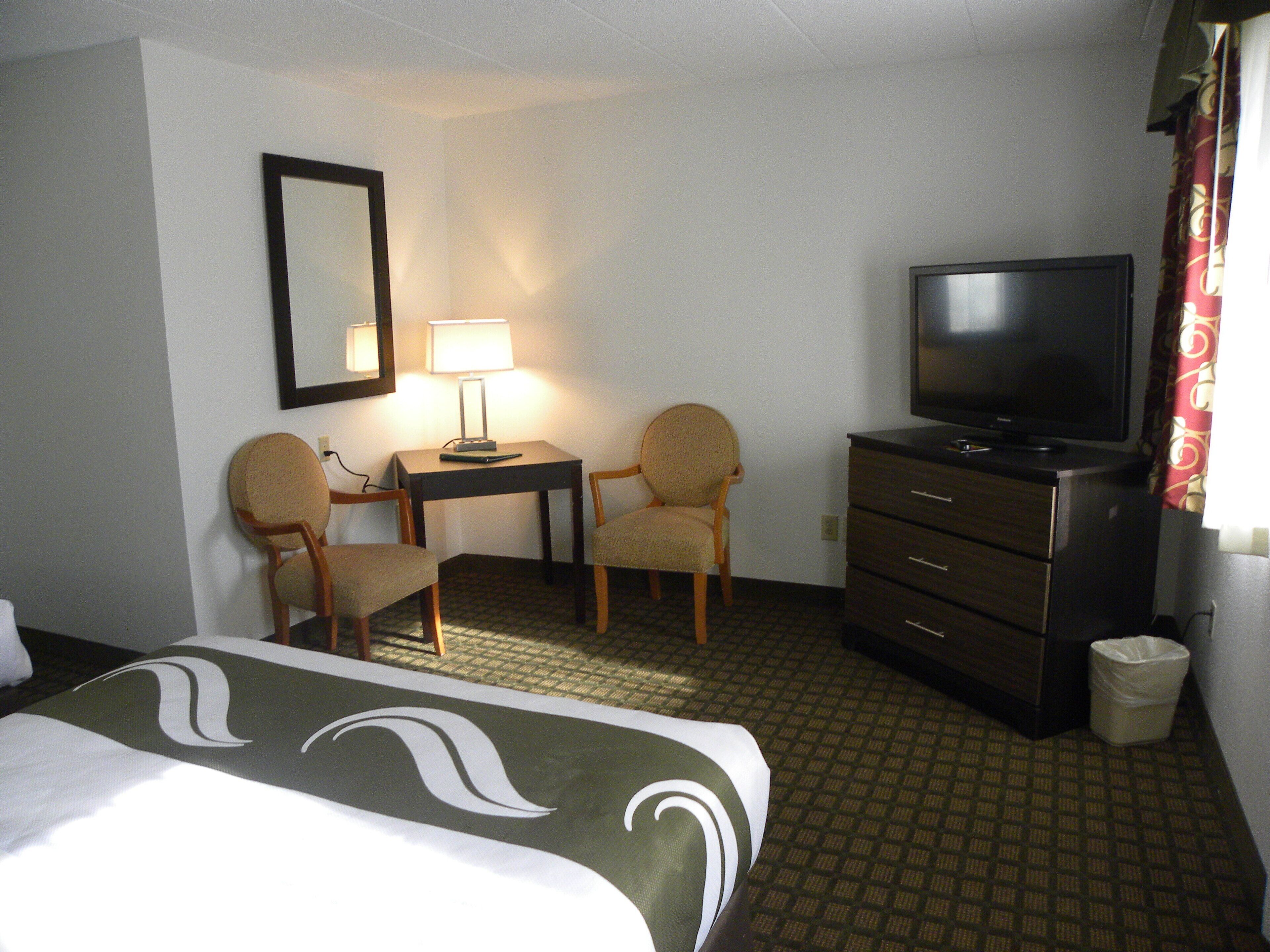 Quarto de hotel com cama, duas cadeiras, escrivaninha com luminária, televisão e cômoda.