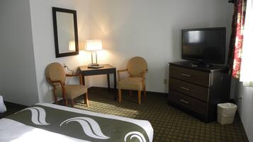 Quarto de hotel com cama, duas cadeiras, escrivaninha com luminária, televisão e cômoda.