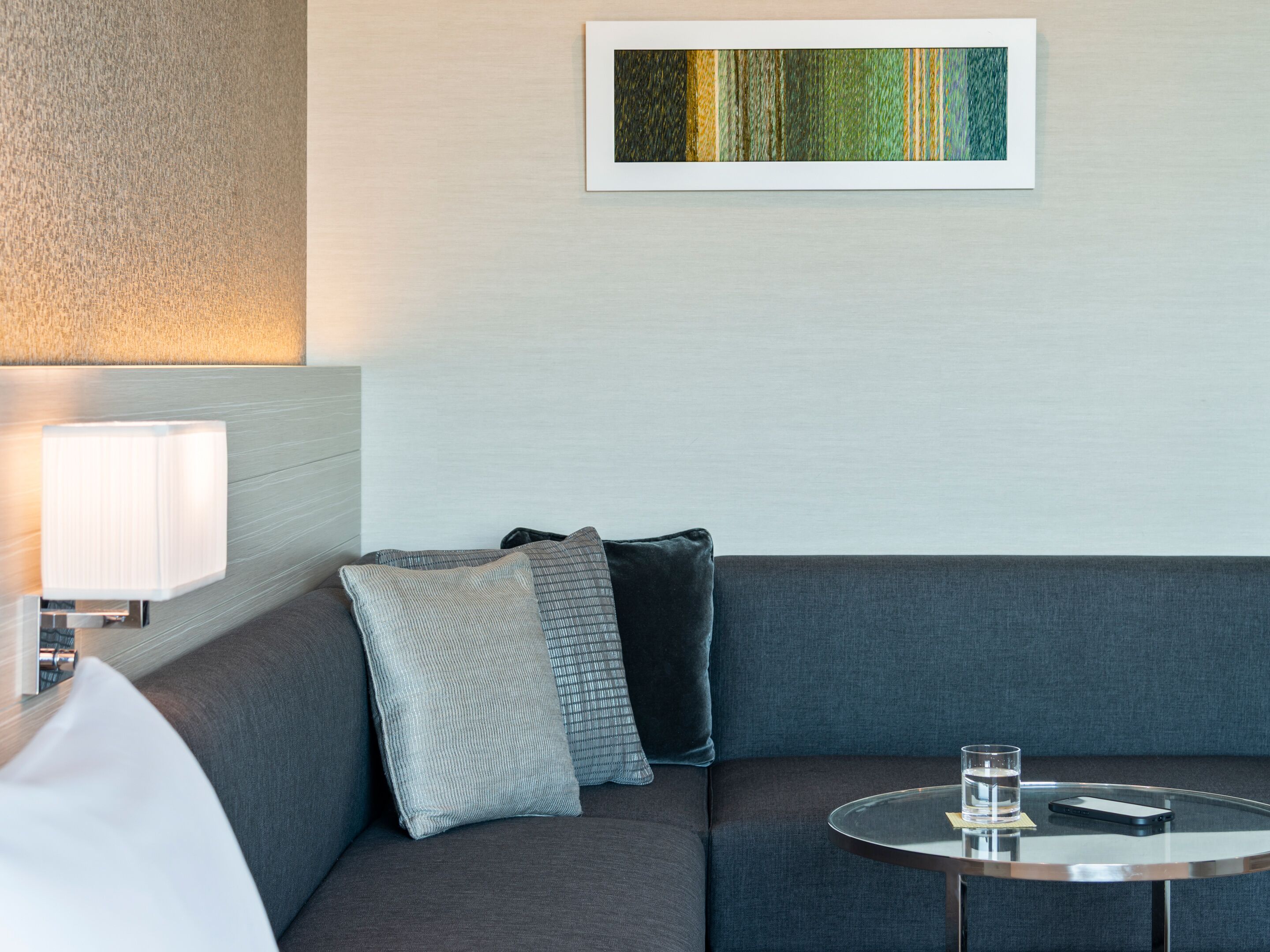 Quarto de hotel moderno com um sofá cinzento, uma mesa de vidro redonda, uma pintura abstrata emoldurada e uma luminária fixada na parede.