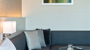 Quarto de hotel moderno com um sofá cinzento, uma mesa de vidro redonda, uma pintura abstrata emoldurada e uma luminária fixada na parede.