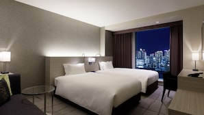 Quarto de hotel com duas camas, uma escrivaninha, uma cadeira, uma mesinha e vista para o horizonte urbano à noite.