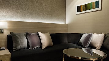 Quarto de hotel moderno com um sofá escuro, uma mesa de centro de vidro, um abajur e um quadro na parede.