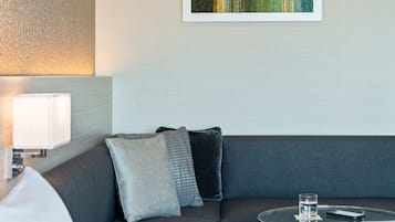 Quarto de hotel moderno com um sofá cinzento, uma mesa de centro de vidro, uma pintura abstrata emoldurada e uma luminária fixada na parede.
