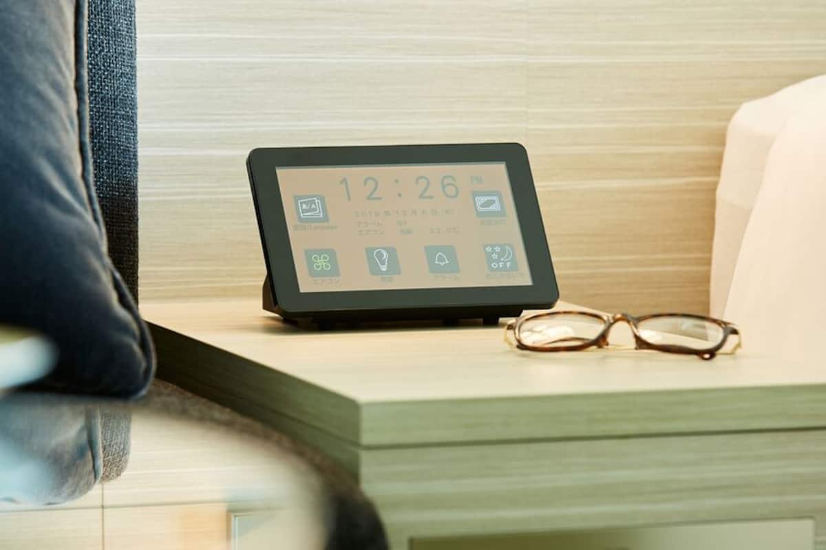 Uma mesa de cabeceira com um relógio digital marcando 12:26, um par de óculos e um travesseiro.