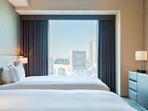 Quarto de hotel com cama grande, vista para a cidade, escrivaninha com abajur e cômoda.