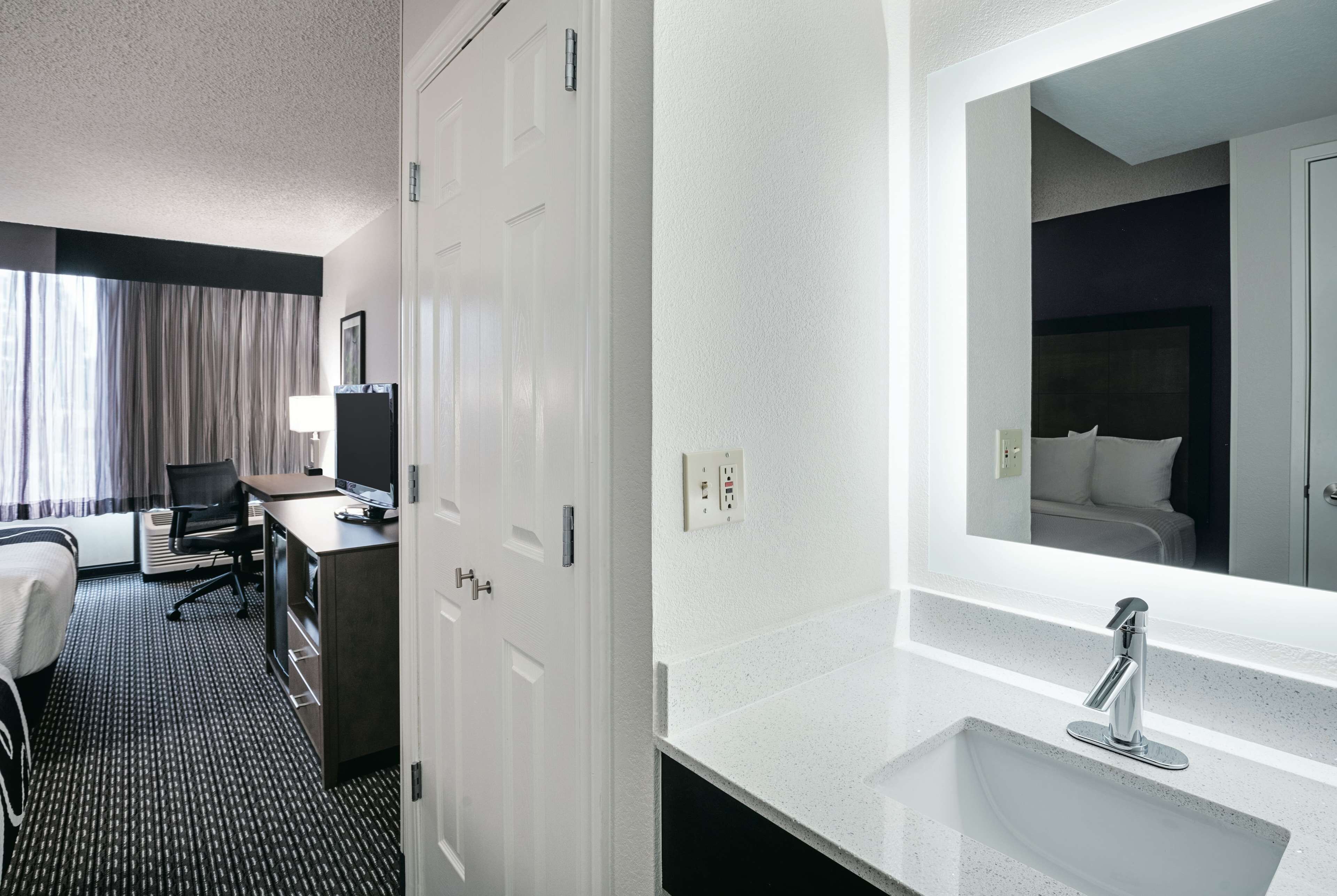 Quarto de hotel com cama, escrivaninha com cadeira, televisão, abajur e banheiro com pia e espelho.