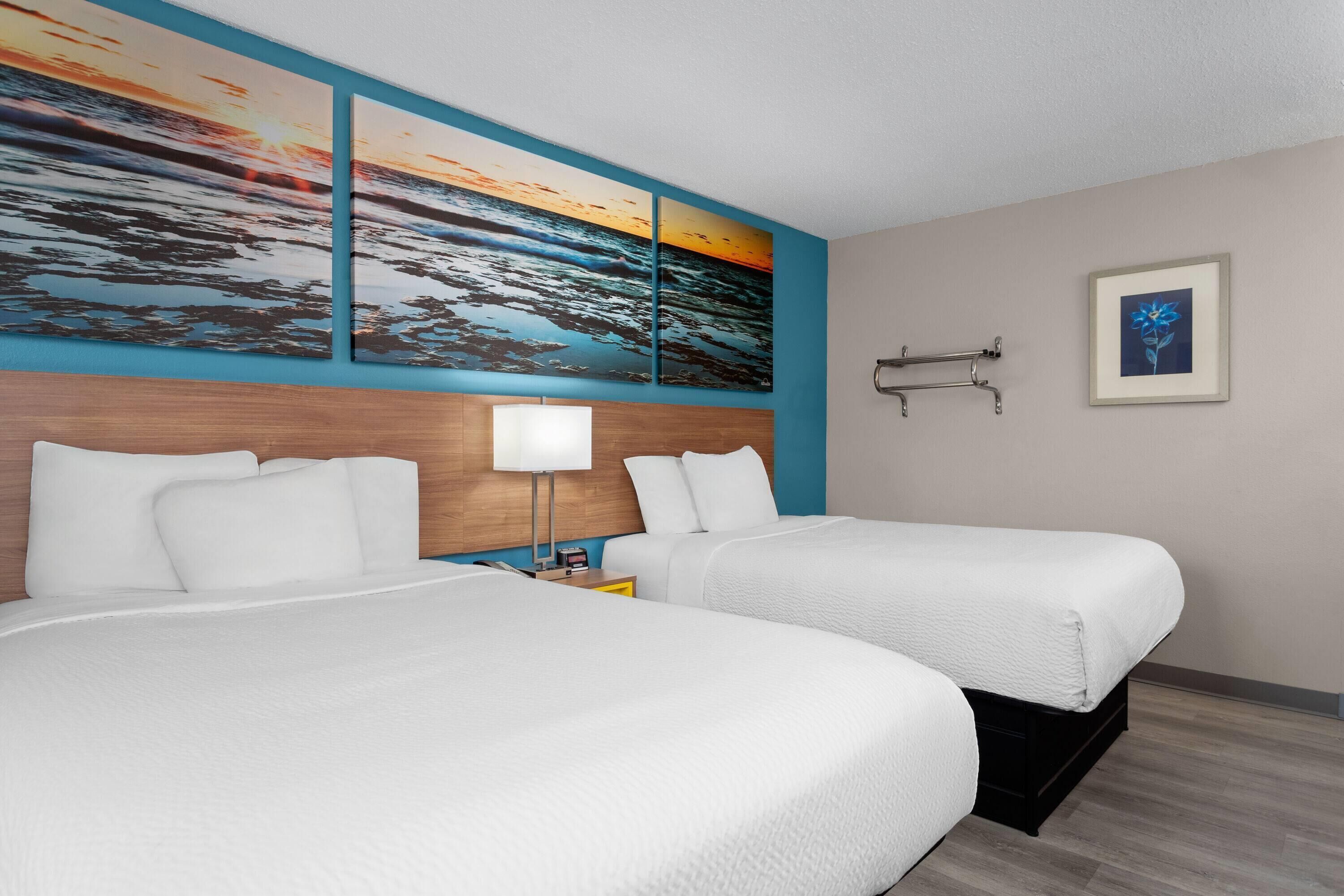 Quarto de hotel com duas camas, uma cabeceira de madeira, uma mesa de cabeceira com abajur, um quadro na parede e vista para o oceano ao pôr do sol.