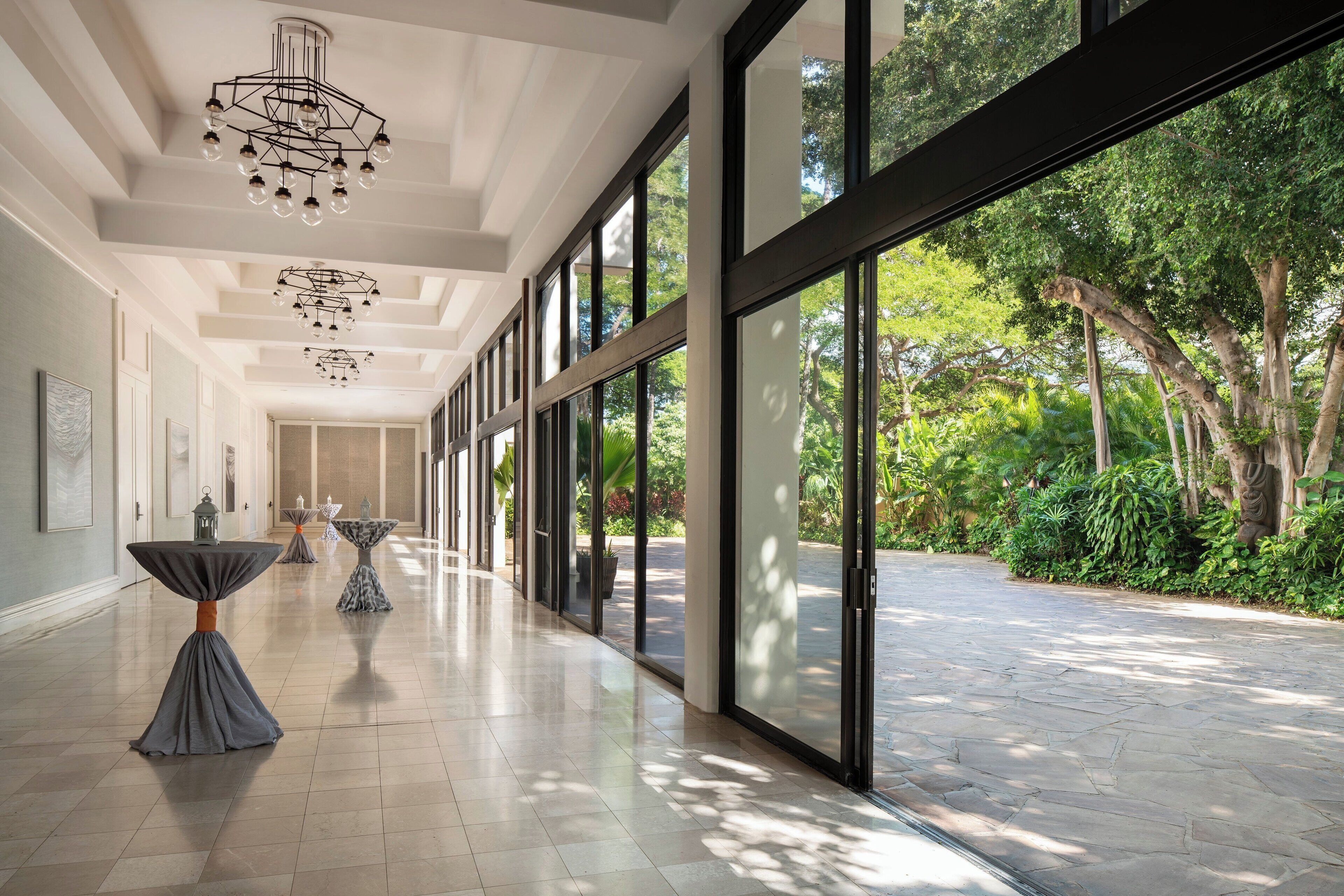Um corredor amplo com grandes janelas, lustres modernos e vista para um jardim exuberante.
