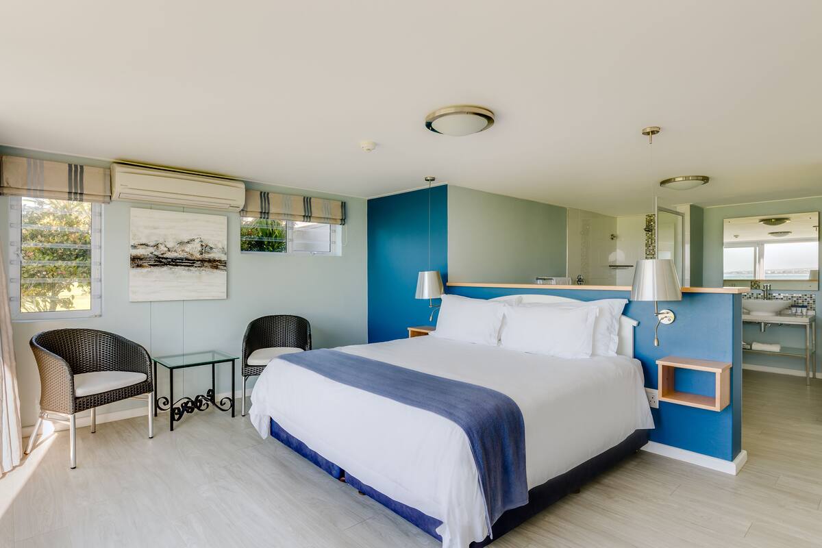 Quarto de hotel moderno com uma cama grande, área de estar com uma poltrona e mesa, banheiro com espelho e pia, e vista para a cidade.
