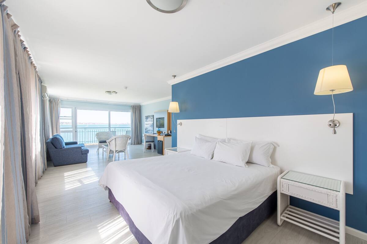 Quarto de hotel com cama grande, escrivaninha, cadeira e vista para o mar.