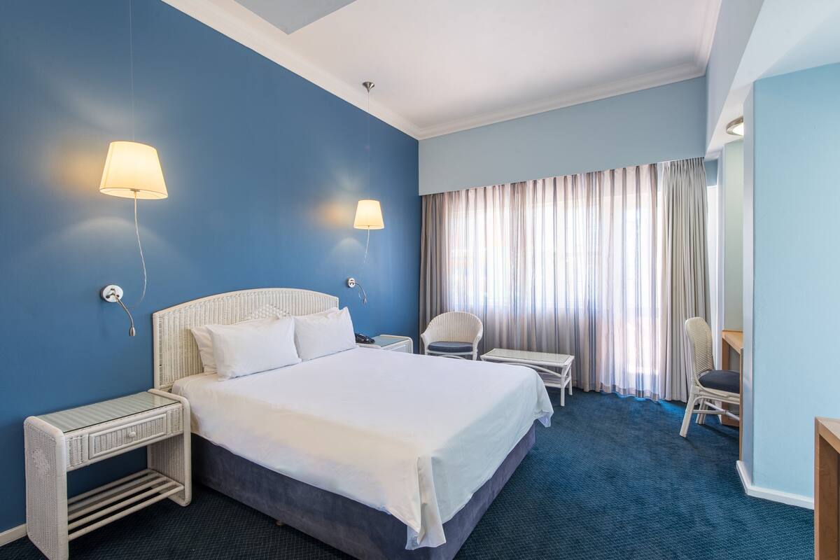 Quarto de hotel com uma cama grande, duas mesas de cabeceira, uma cadeira, uma escrivaninha e uma janela com cortinas.