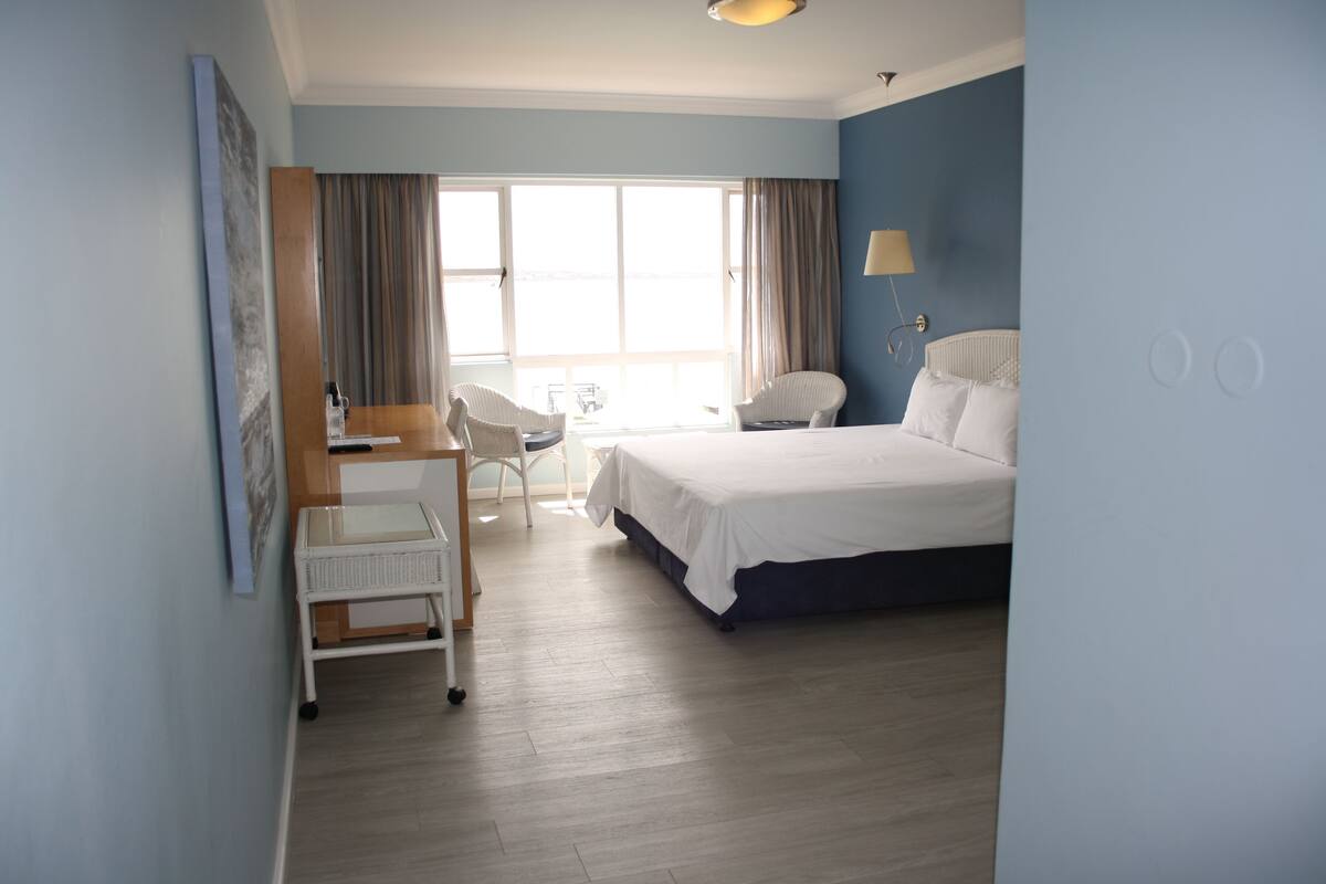 Quarto de hotel com cama, escrivaninha, cadeira e janela com cortinas.