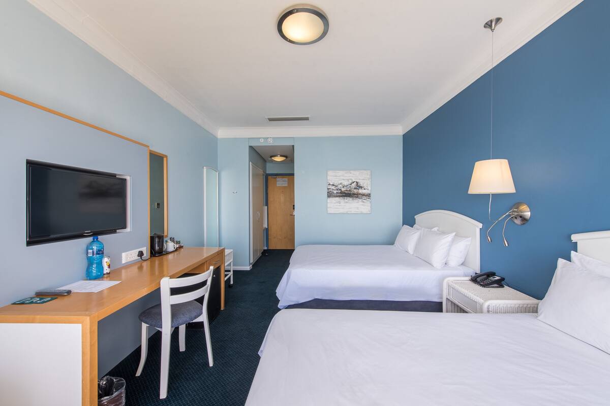 Quarto de hotel com duas camas, uma escrivaninha, uma cadeira, uma televisão e um abajur.