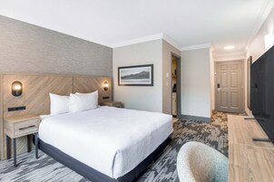 Quarto de hotel com uma cama grande, cabeceira de madeira, mesa de cabeceira, escrivaninha, cadeira, televisão de tela plana e quadro na parede.