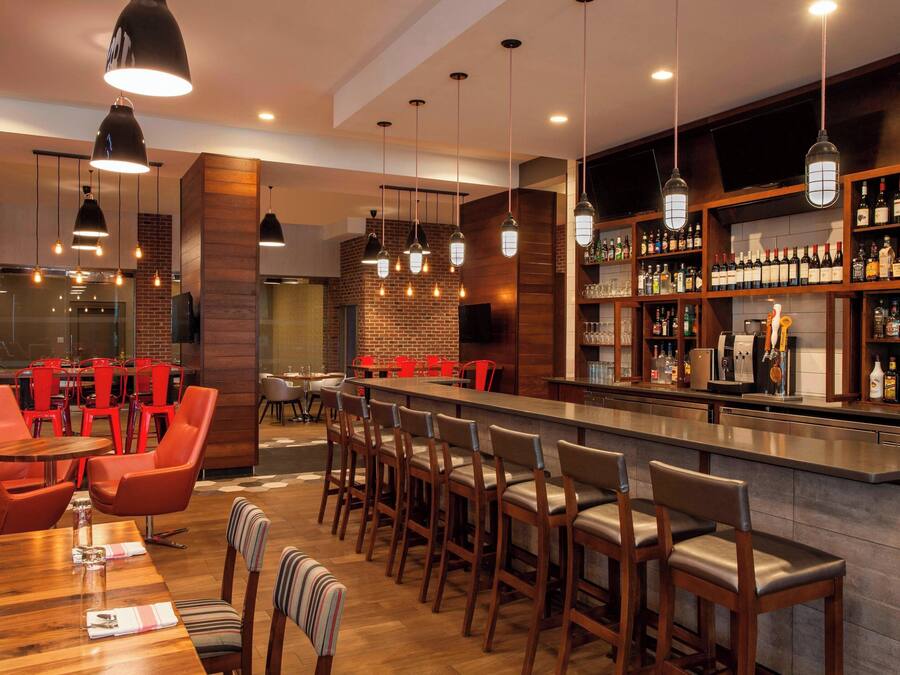 Uma área de bar com várias opções de assento, incluindo banquetas e cadeiras vermelhas, e uma prateleira de bebidas bem abastecida.