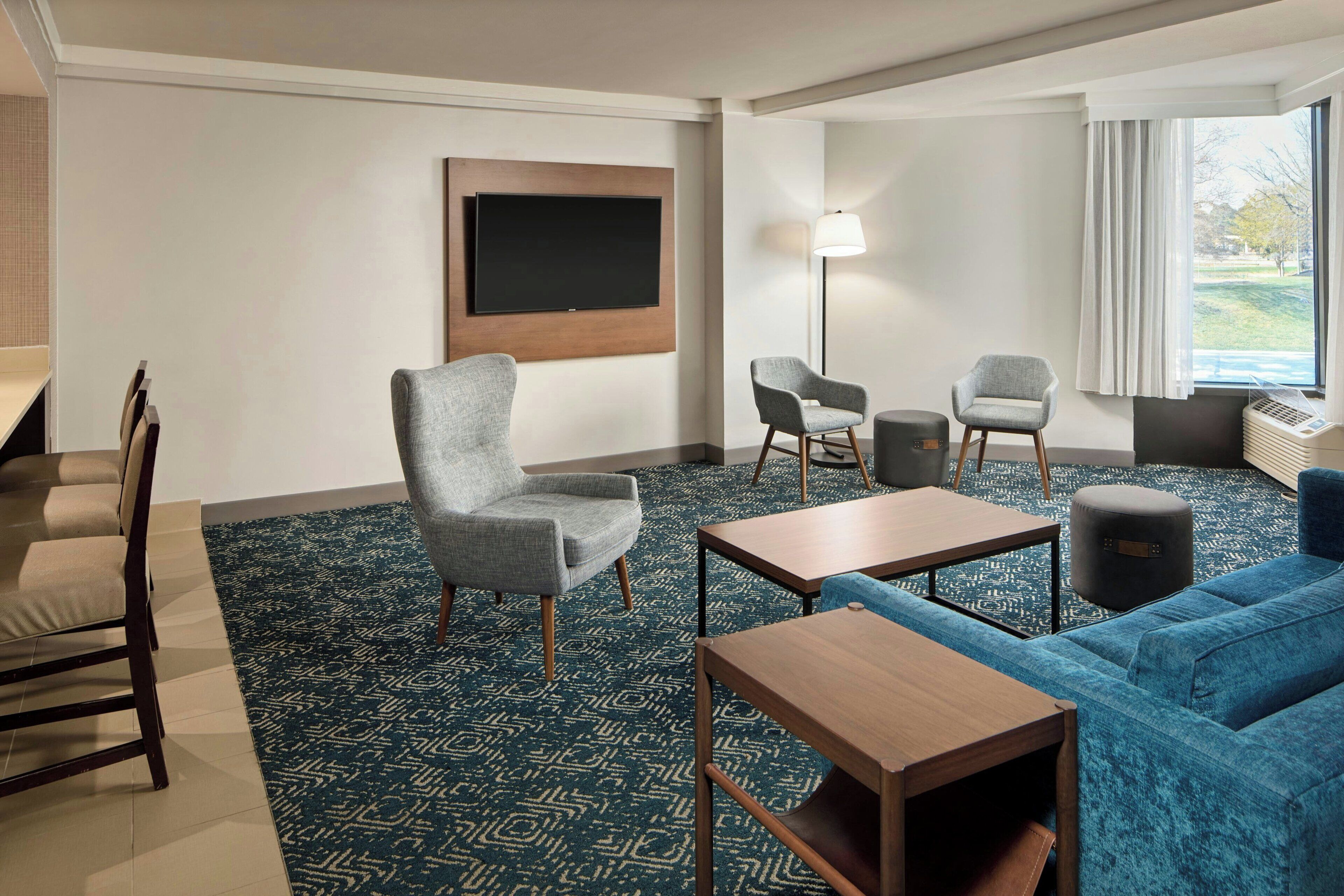 Quarto de hotel com uma janela grande, televisão de tela plana presa na parede, carpete azul estampado e sofá azul.