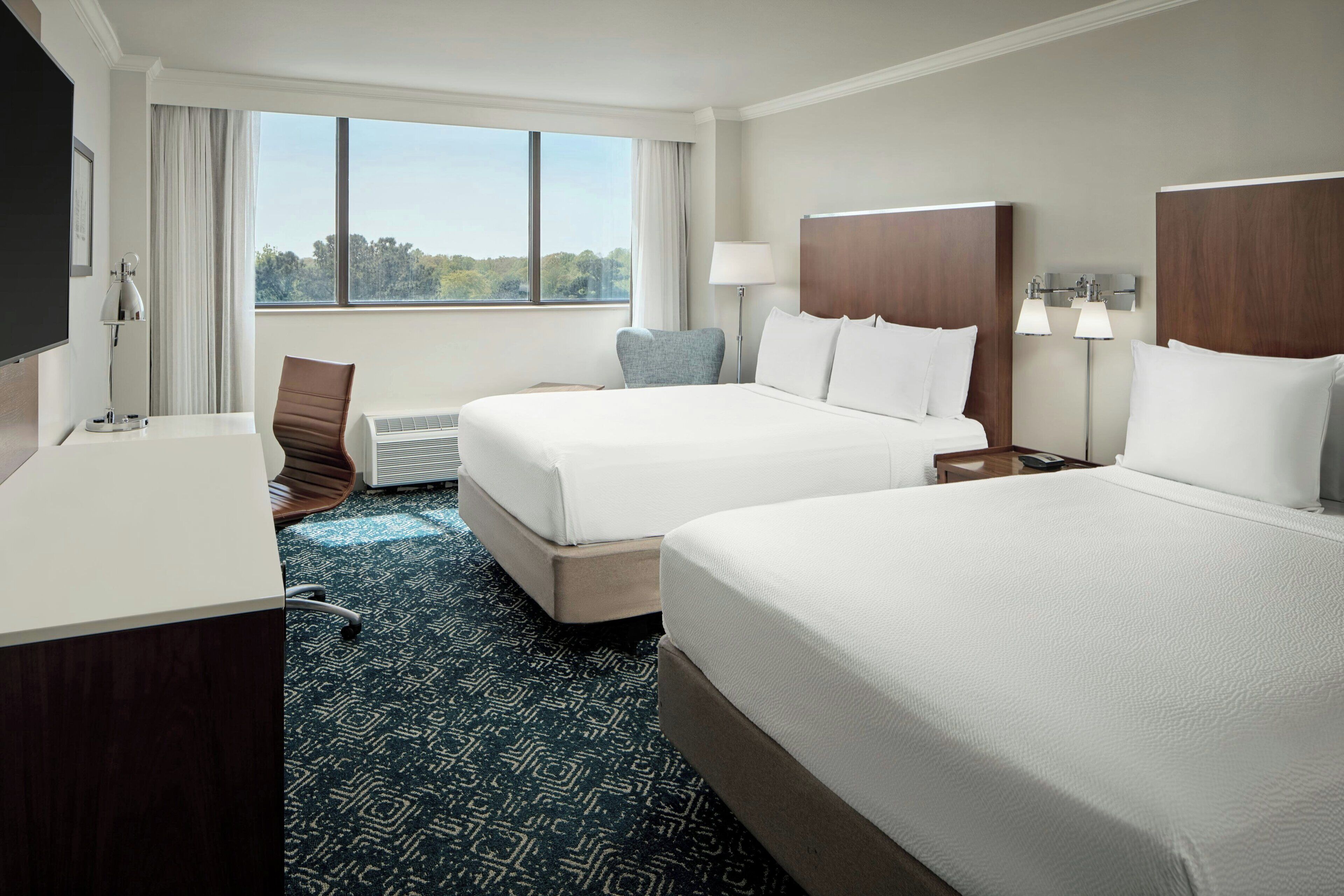 Quarto de hotel com duas camas, uma escrivaninha, uma cadeira, um abajur e janela com vista para árvores.