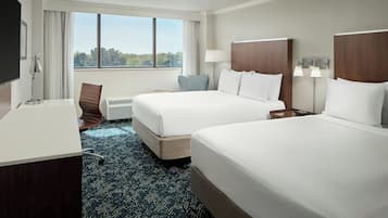 Quarto de hotel com duas camas, uma escrivaninha, uma cadeira, um abajur e janela com vista para árvores.