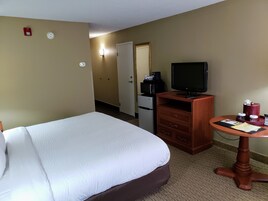 Quarto de hotel com uma cama, uma televisão sobre um suporte de madeira, um frigorífico e uma pequena mesa com um tabuleiro.