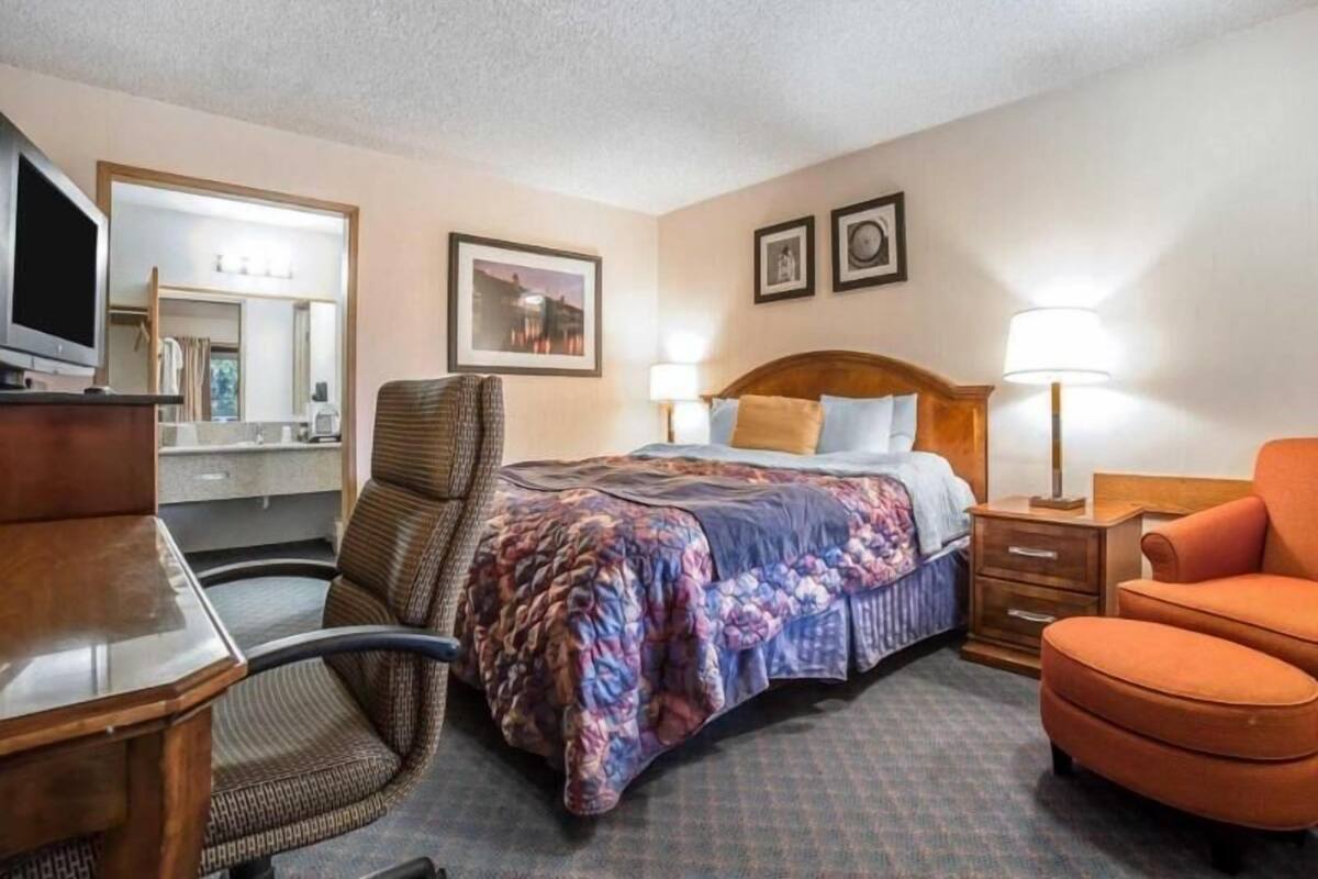 Quarto de hotel com cama, escrivaninha, cadeira, televisão e luminária.