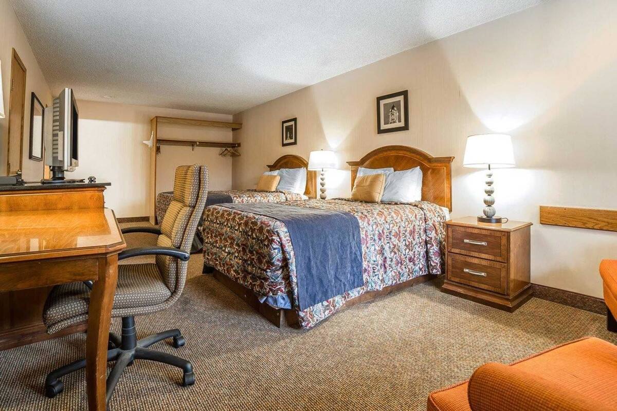 Quarto de hotel com cama, escrivaninha, cadeira e uma pequena mesa com abajur.