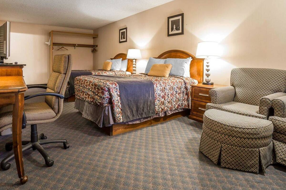 Quarto de hotel com uma cama grande, duas mesas de cabeceira com luminárias, uma escrivaninha de madeira, uma cadeira e um tapete estampado.