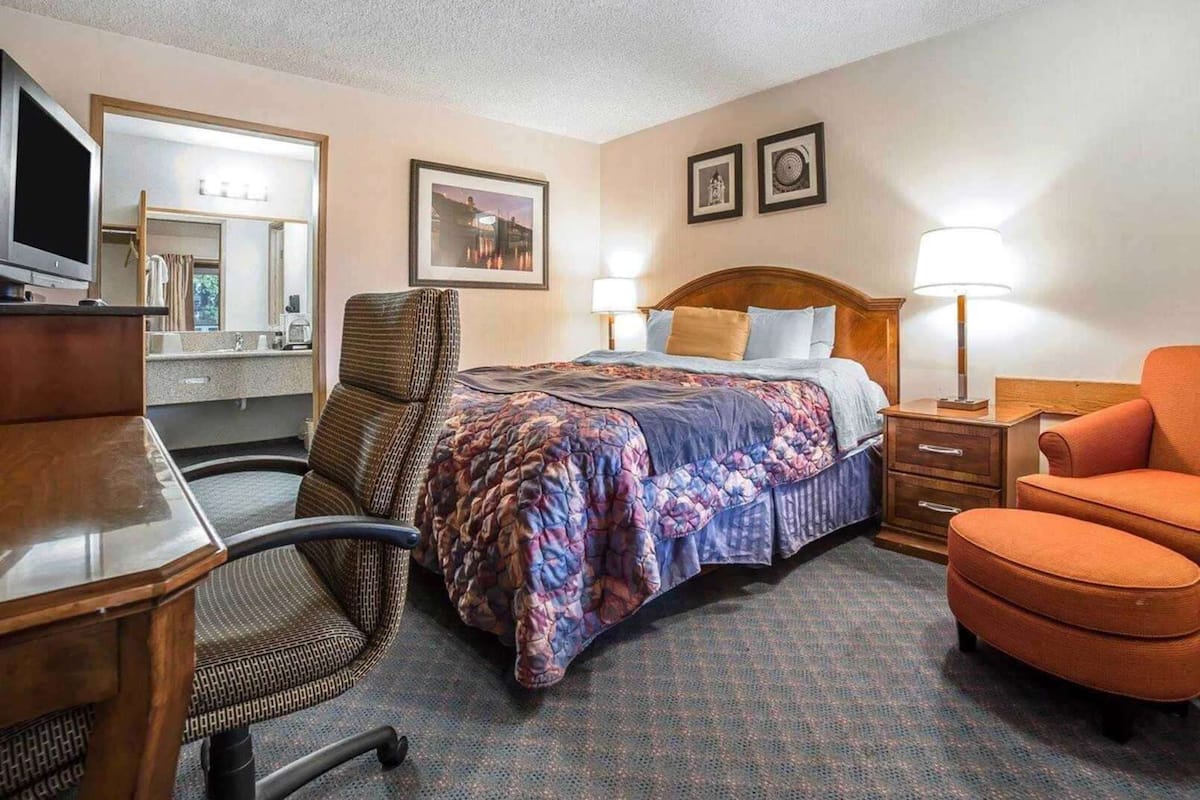 Quarto de hotel com uma cama, uma escrivaninha com televisão, uma cadeira, um abajur e uma poltrona laranja.