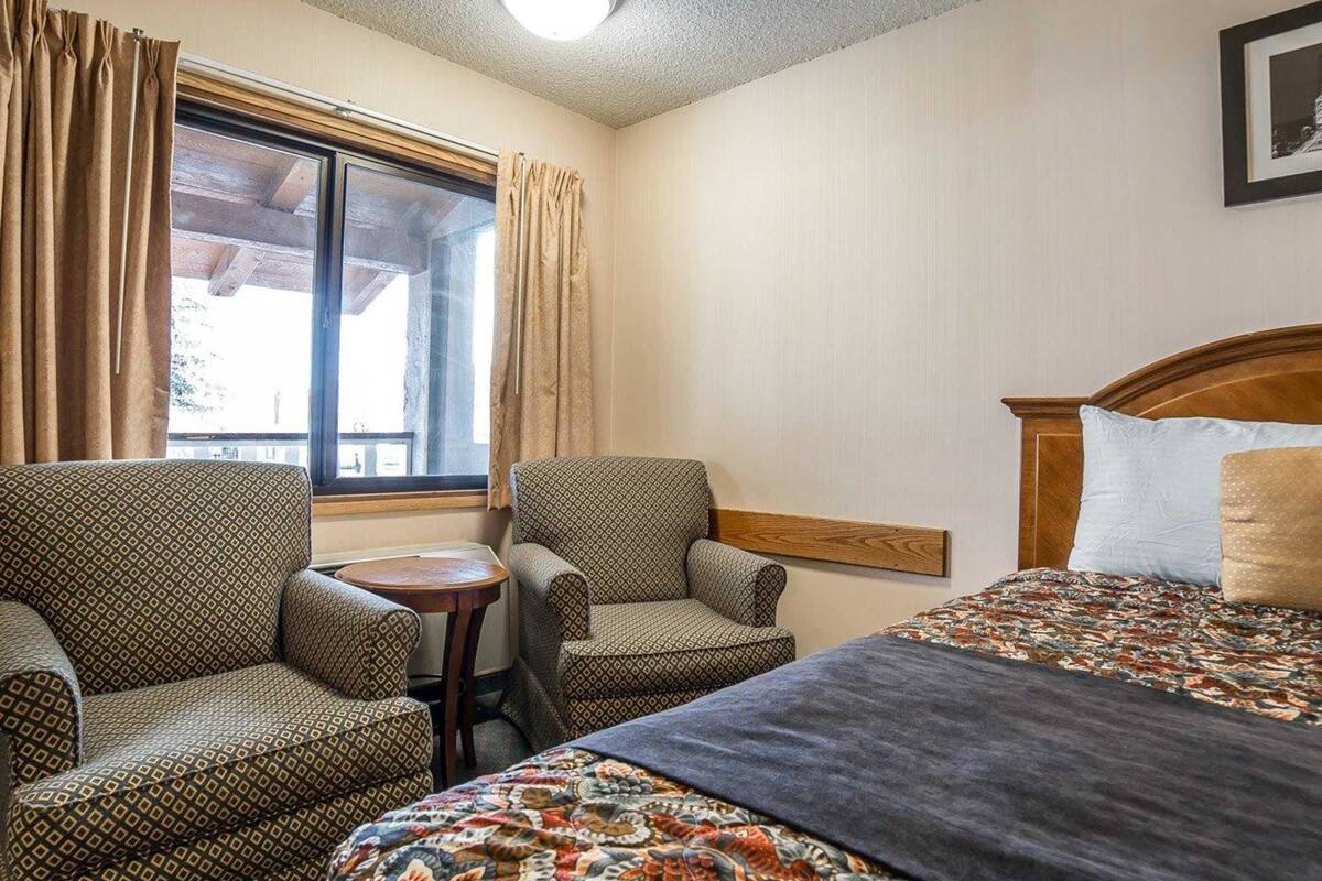 Quarto de hotel com uma cama, duas poltronas estampadas, uma mesinha e uma janela com cortinas.
