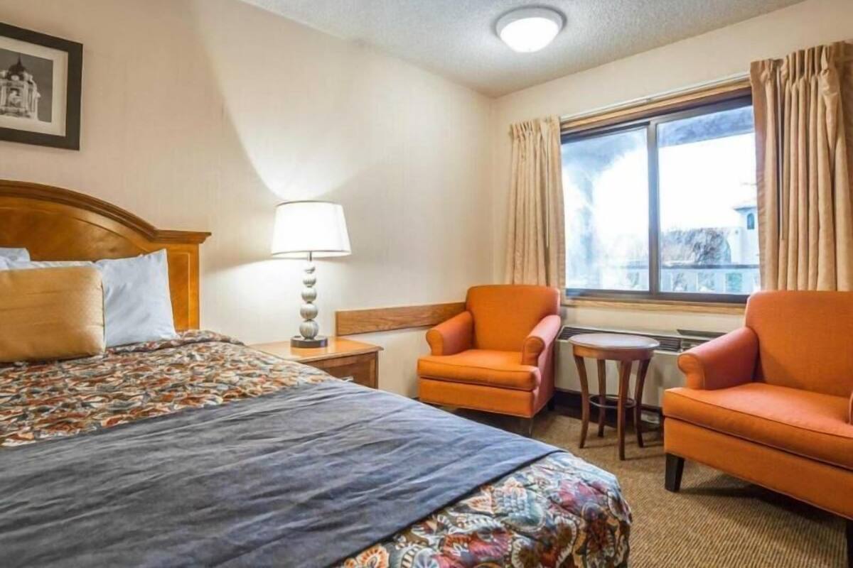 Quarto de hotel com cama, duas poltronas, uma mesinha, abajur, janela com cortinas e um quadro na parede.