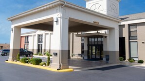 Entrada de hotel com área de espera coberta, torre do relógio e estacionamento.