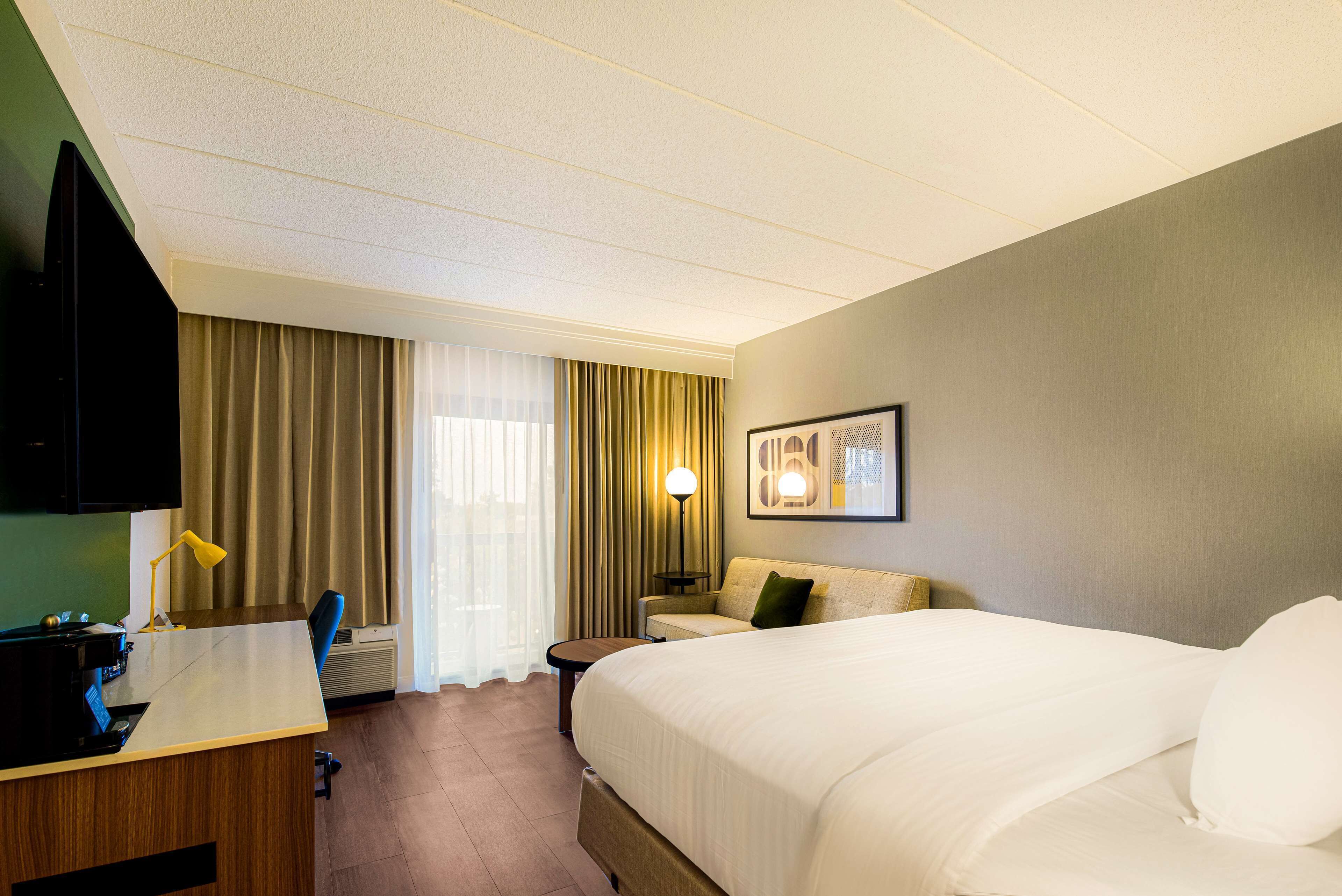 Quarto de hotel com cama, escrivaninha, cadeira, televisão, luminária e janela com cortinas.