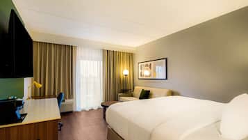 Quarto de hotel com cama, escrivaninha, cadeira, televisão, luminária e janela com cortinas.