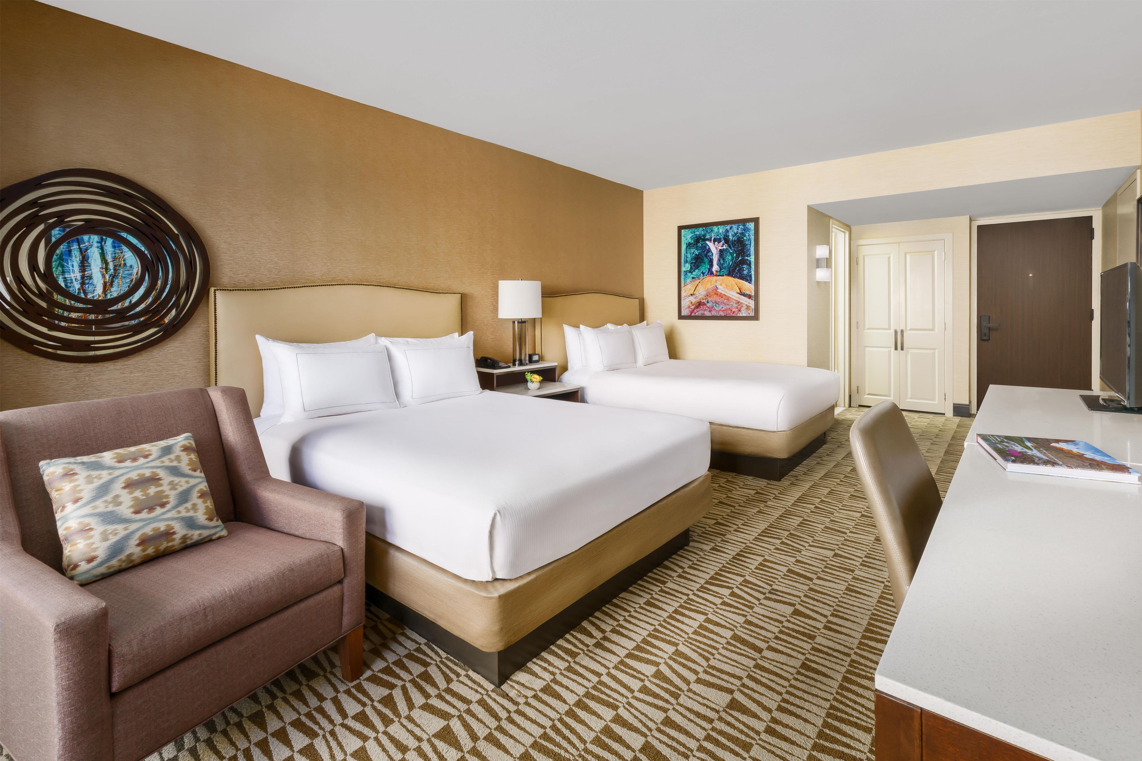 Quarto de hotel com uma cama grande, uma cama menor, uma escrivaninha, uma cadeira, uma mesinha de cabeceira, um abajur, um quadro e uma porta.