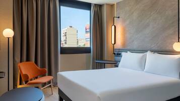 Quarto de hotel com cama, cadeira, mesa pequena, abajur e vista para edifícios do lado de fora da janela.