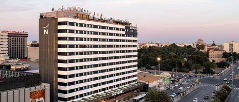 Um hotel de vários andares com um letreiro chamativo 'NOVOTEL', um terraço no telhado e vista para a paisagem urbana ao redor.