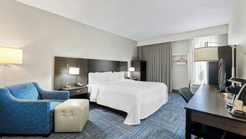 Quarto de hotel com cama, escrivaninha, cadeira, abajur, mesa de cabeceira e televisão.