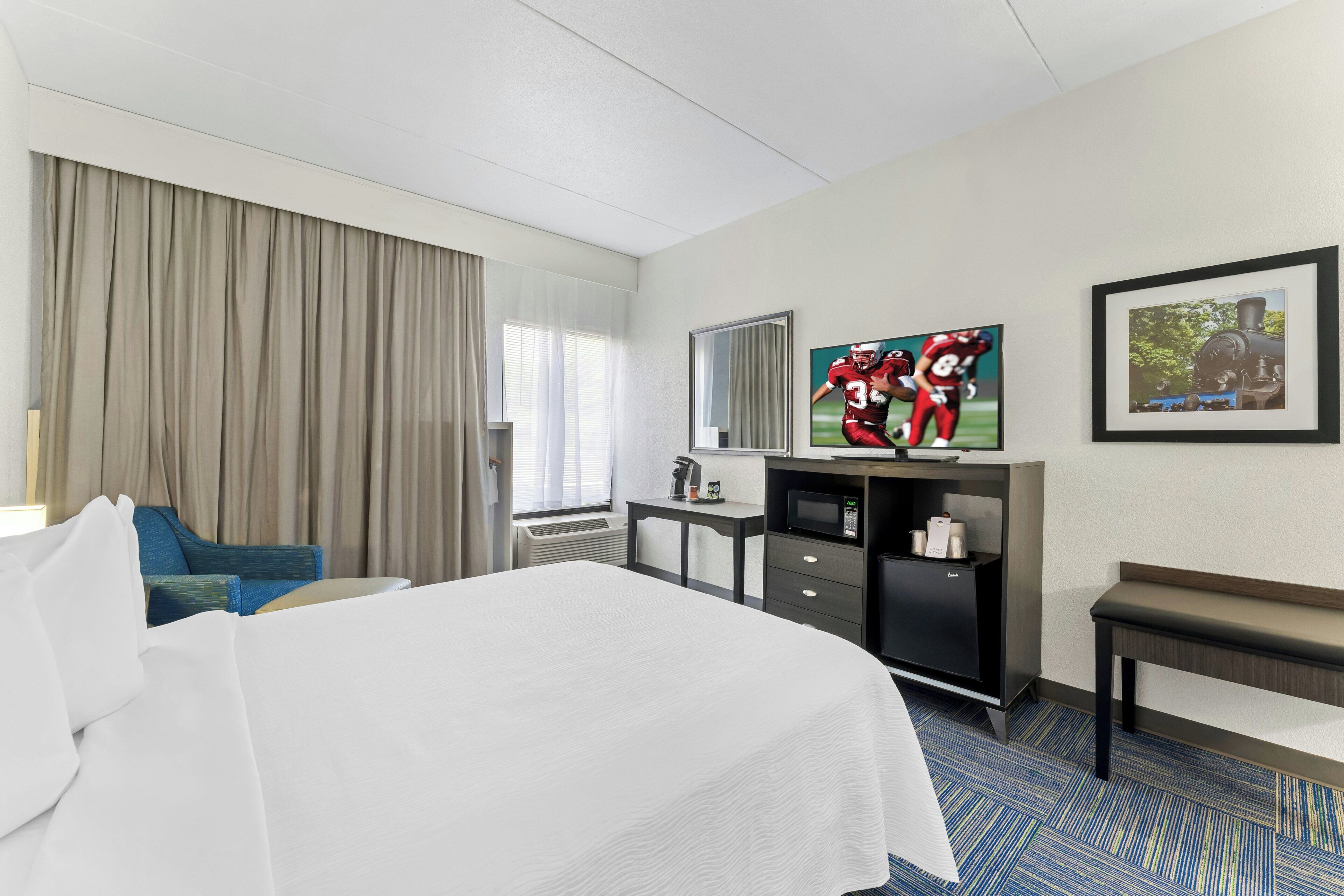 Quarto de hotel com uma cama grande, uma escrivaninha com computador, televisão, espelho e um quadro na parede.
