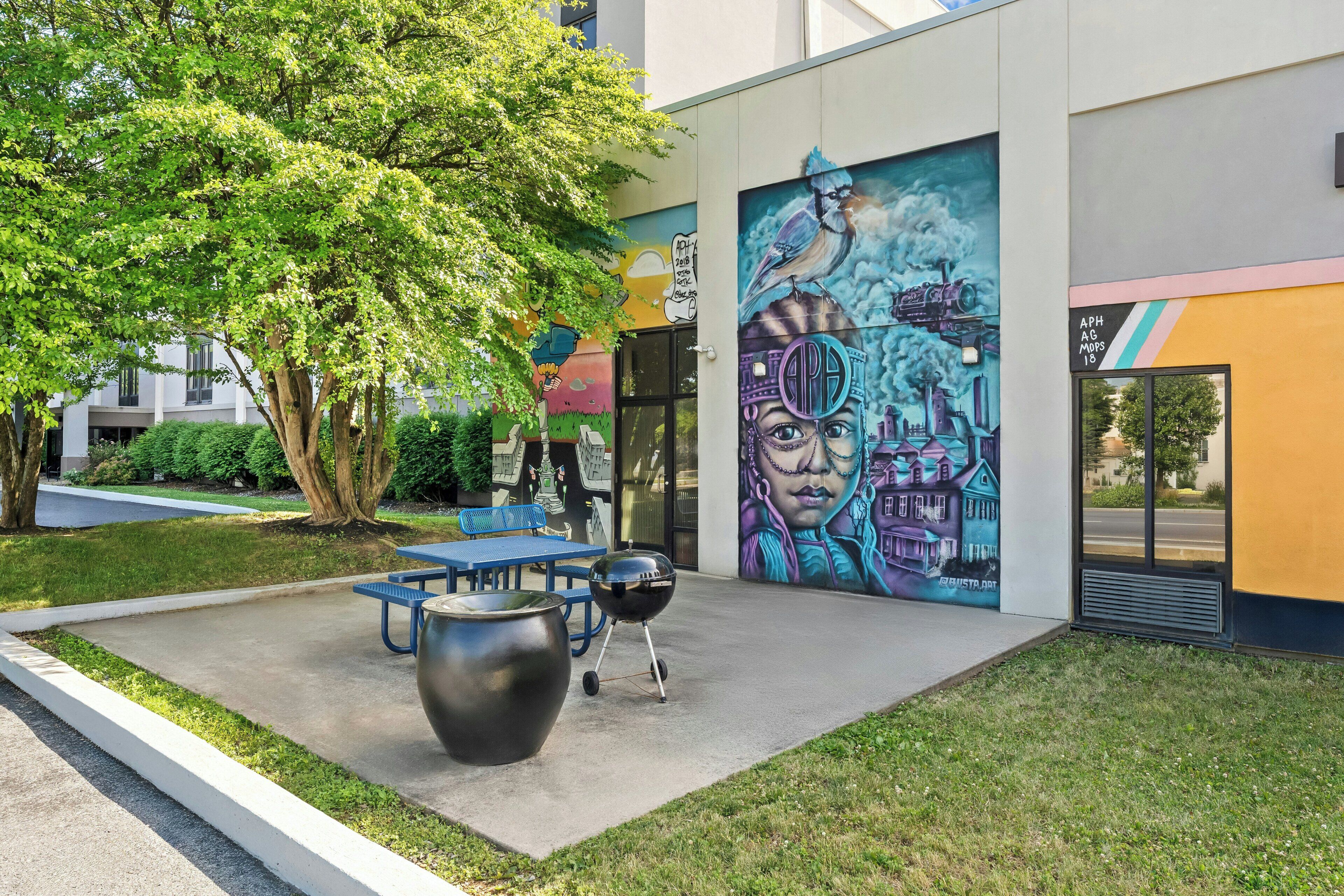 Uma área externa com mesas, churrasqueira, um mural na fachada e uma árvore.