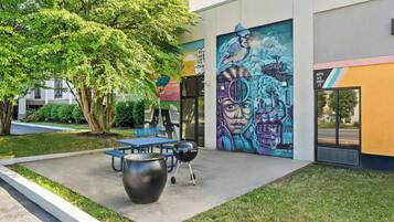 Uma área externa com mesas, churrasqueira, um mural na fachada e uma árvore.
