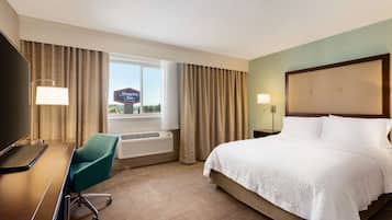 Quarto de hotel com uma cama, uma escrivaninha com cadeira, uma televisão e uma janela com cortinas.
